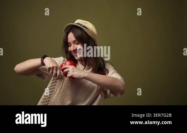 Young smiling woman in a straw hat putting fresh red bell pepper into ...
