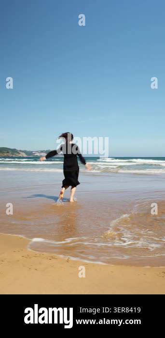 A carefree woman in a black dress dances and spins barefoot on a sandy ...