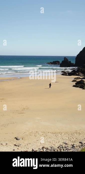 A lone woman in a black dress walks along a vast, sunny beach towards ...
