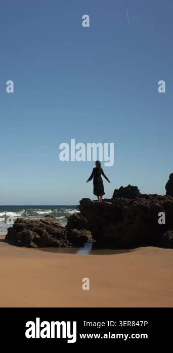 A woman in a dark dress stands on a rock at a sandy beach, looking out ...