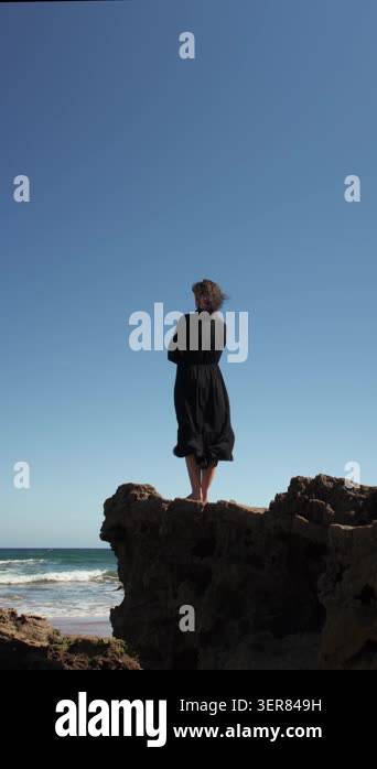 A woman in a black dress stands on a rocky cliff overlooking the ocean ...
