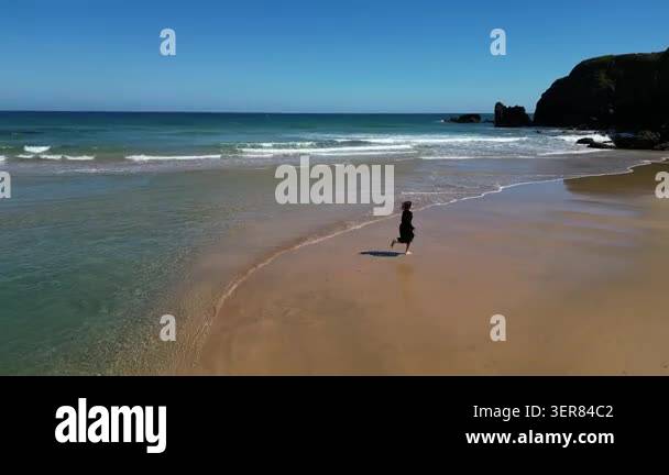 Aerial view of a young woman in a black dress running barefoot along ...