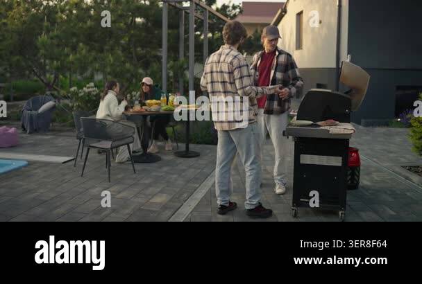 Two men stand near a barbecue grill in a residential backyard while ...