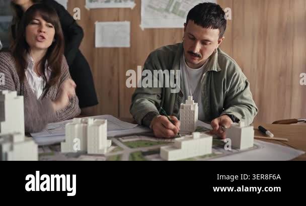 A man in the foreground examines and marks a building model on a table ...
