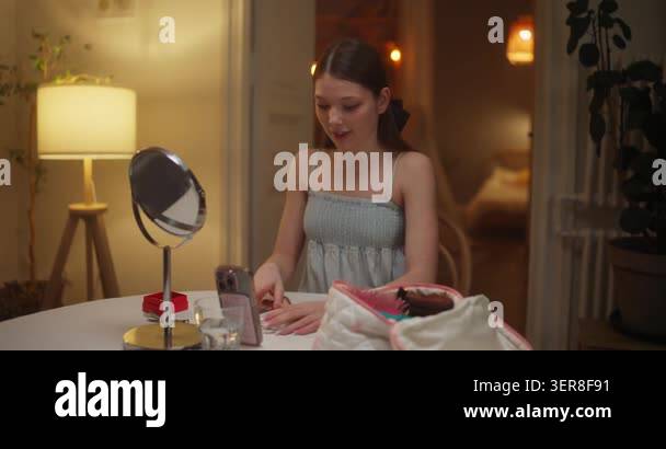 Young girl sits at a table indoors, preparing makeup beside a mirror in ...