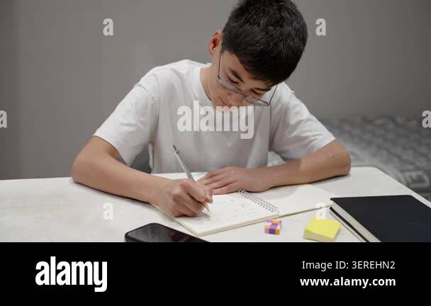 Front view of teenage student in glasses writing in notebook at home ...