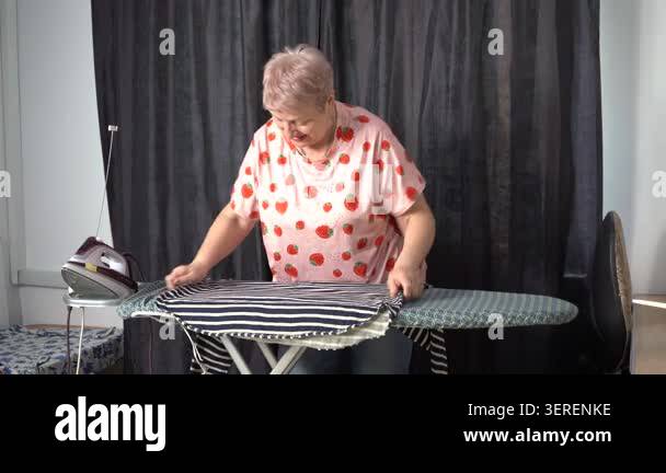 Smiling senior elderly woman ironing clothes with an iron on a board ...