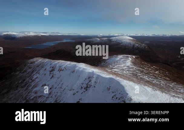 Aerial drone flyover of a snow-covered ridge with a distant loch and ...
