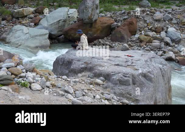 A person sits on a rock beside a rushing river during a trek in the ...
