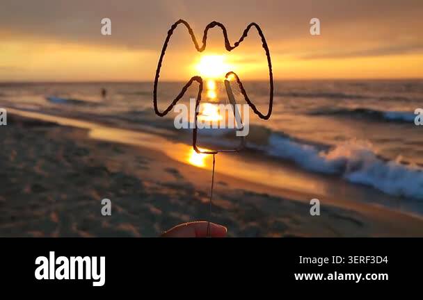Burning sparkler in shape of angel against backdrop of sea at sunrise ...