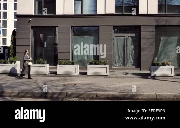 Wide shot of a stylish woman in a grey coat walking past modern ...