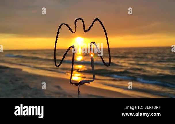 Burning sparkler in shape of angel against backdrop of sea at sunrise ...
