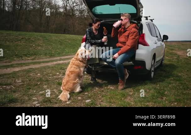 A couple relaxes at their car while eating snacks and drinking coffee ...