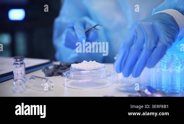 Lab worker uses metal tweezers to place small white tablets into tiny ...