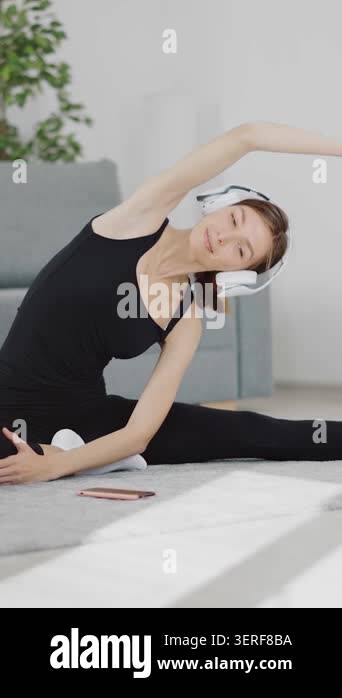 A woman stretches on a mat in a bright room with plants and headphones ...