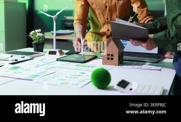 Team reviewing eco home plans at a desk with a model house, wind ...
