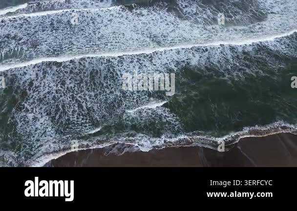 Aerial view of ocean waves crashing on a dark sandy beach, showcasing ...