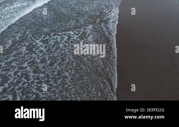 Aerial view of ocean waves crashing on a dark sandy beach, showcasing ...