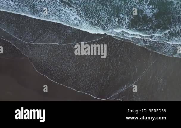Aerial view of ocean waves crashing on a dark sandy beach, showcasing ...