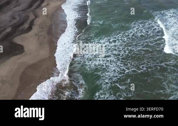 Aerial view of ocean waves crashing on a sandy beach with dark pebbles ...