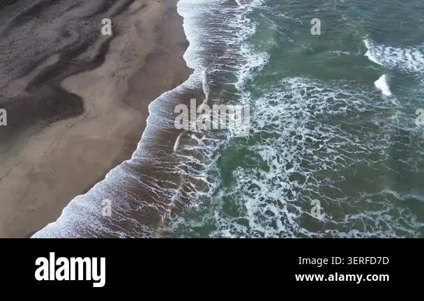 Aerial view of ocean waves crashing on a sandy beach with dark sand ...
