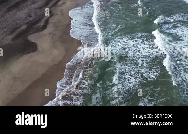 Aerial view of ocean waves crashing on a sandy beach with dark pebbles ...