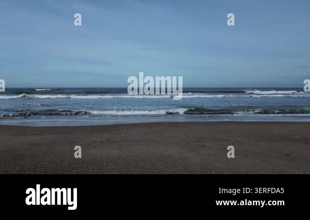 Wide shot of ocean waves crashing on a sandy beach under a cloudy sky ...