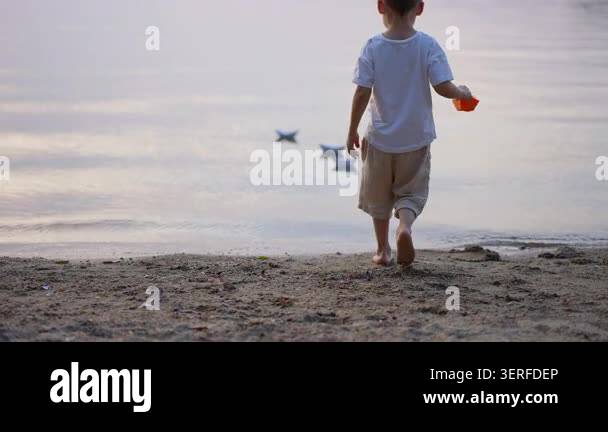 Barefoot child walking toward paper boats floating on lake. Boy holds ...