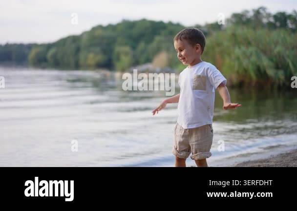 Smiling boy walking along riverbank with arms outstretched in summer ...