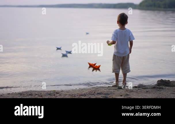 Boy standing in shallow water watching paper boats on lake. Rear view ...