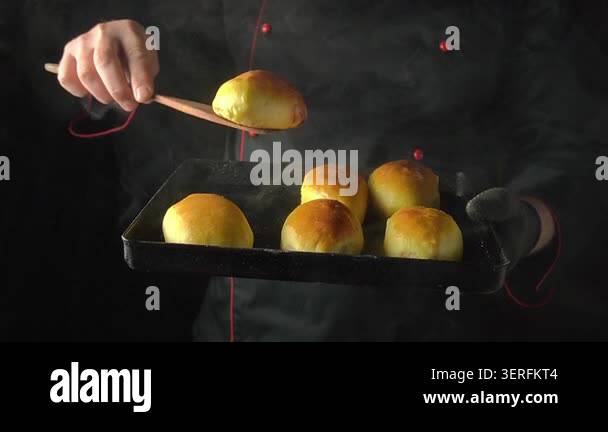 Chef in a black uniform holds a tray of golden rolls, lifting one with ...