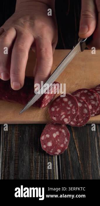 Hand positioned over a cured sausage on a wooden cutting board, slicing ...