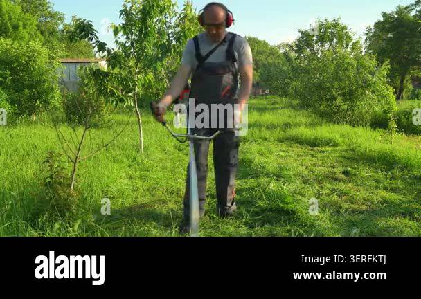 Worker in gray overalls and safety gear operates a grass trimmer ...