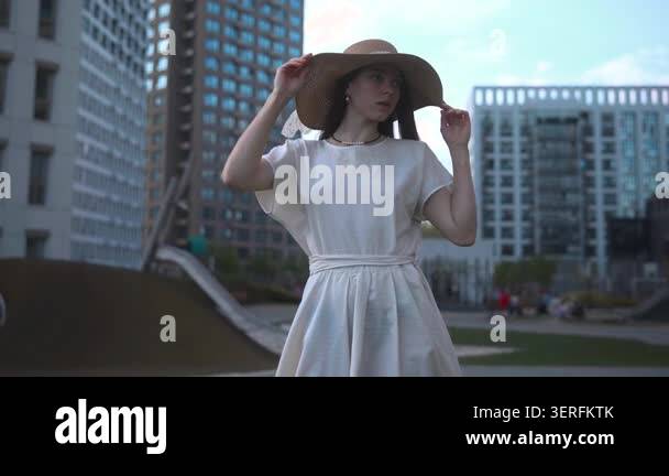 A young woman in a flowing white dress stands in an open urban plaza ...