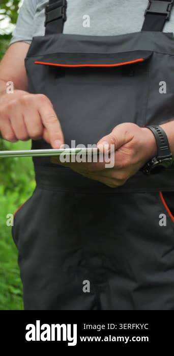 Man in gray shirt and black overalls stands outdoors, using a tablet ...