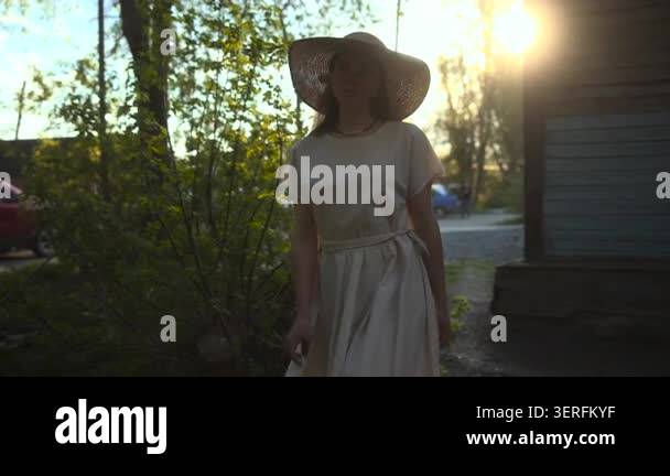 A woman wearing a light dress and wide-brimmed hat stands outdoors near ...
