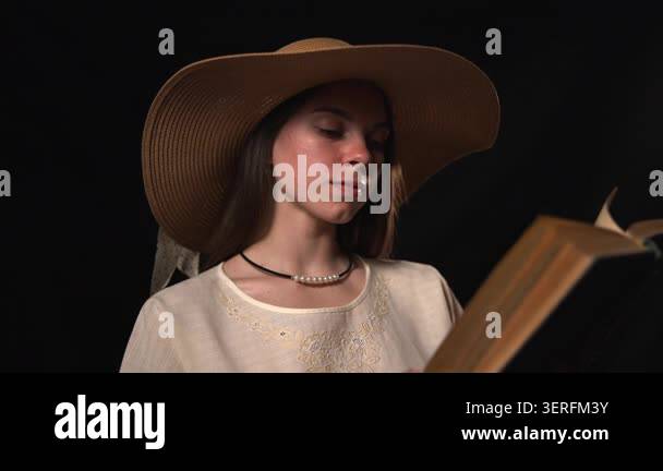 A young woman wearing a wide-brim hat reads an open book against a dark ...