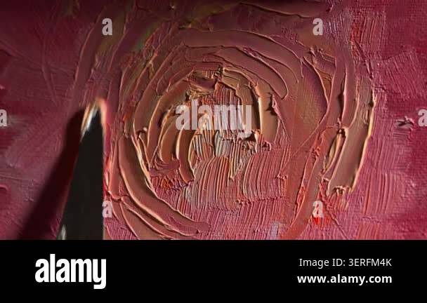 Close-up of a hand using a palette knife to spread thick pink and brown ...