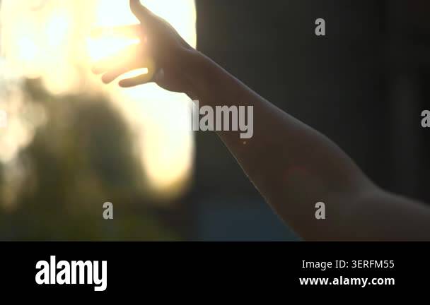A silhouetted arm and hand reach upward toward intense sunlight in a ...