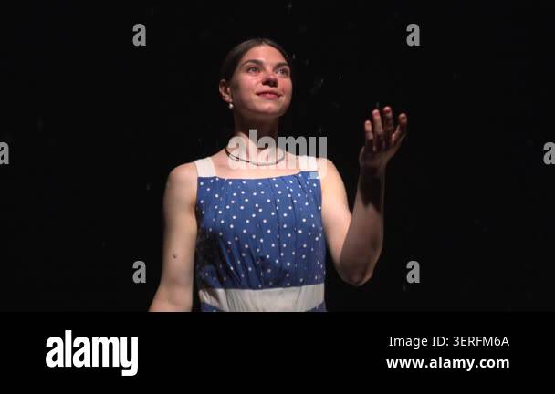 A young woman stands against a dark background, looking upward with one ...