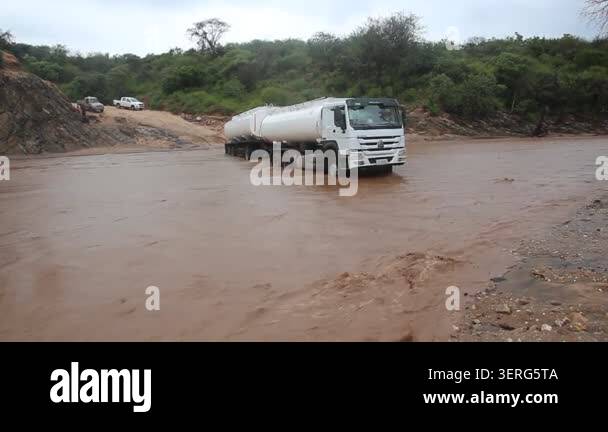 Tanker truck drives through muddy water crossing a riverbed with trees ...