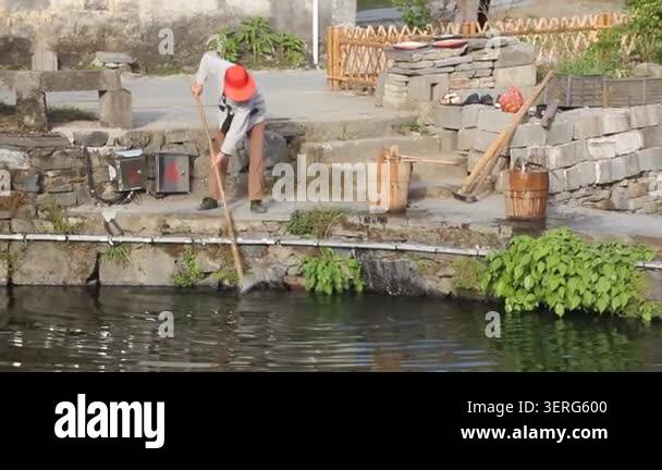 Person wearing a red hat scoops water from a pond using a long handled ...
