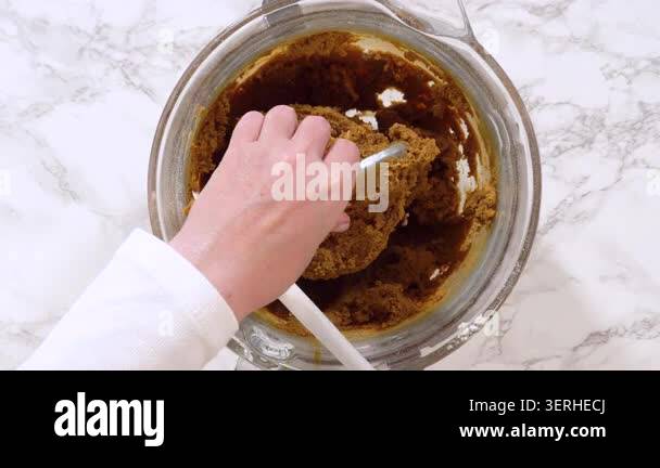 Hands scrape thick brown dough from a stand mixer bowl using a spatula ...