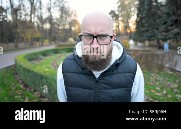 Bald man with glasses and beard stands in lush green maze at park ...