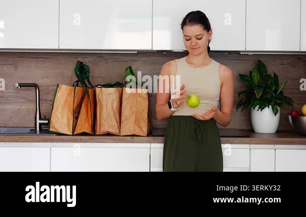 Woman with fresh apple standing near reusable grocery bags in kitchen ...