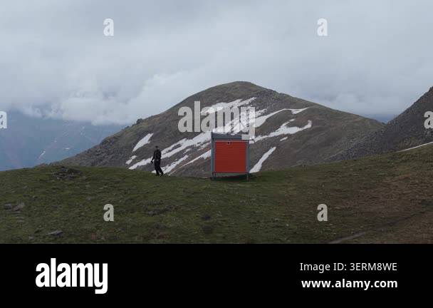 A hiker in dark clothing walks towards an orange shelter on a grassy ...