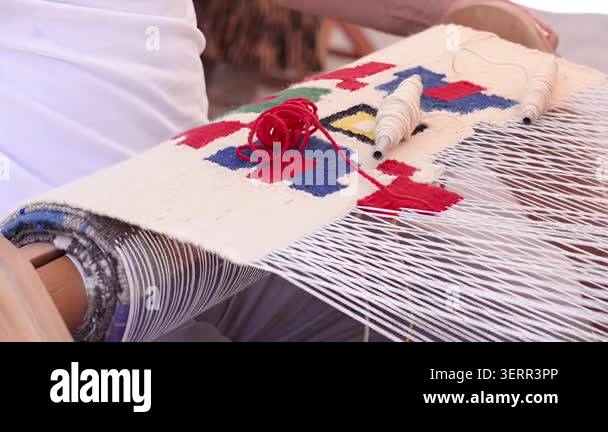 Hands weave a textile with red, blue, green, and white patterns on a ...
