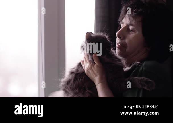 A senior woman gently holds her gray cat near a bright window ...