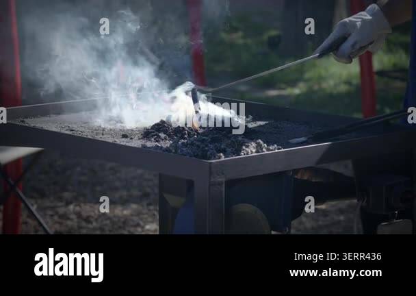 A gloved blacksmith uses long tongs over a coal forge as white smoke ...