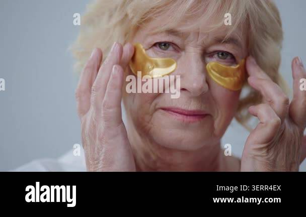 Good-looking elderly woman applying golden eye patches under her eyes ...